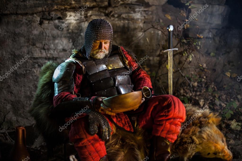 Old medieval King in armor with dish is sitting on furs near the ...