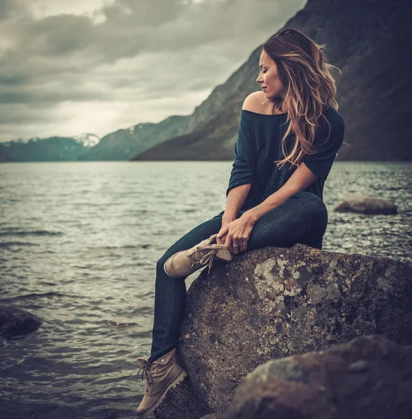 Beautiful woman posing on the shore of a wild lake, with mountains on ...
