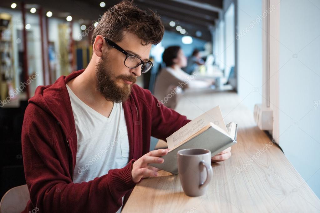 Chico listo en gafas leyendo libro sentado en el escritorio 2022