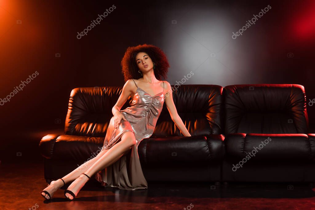 longitud completa de la mujer elegante en vestido sentado en el sof sobre fondo negro 2024