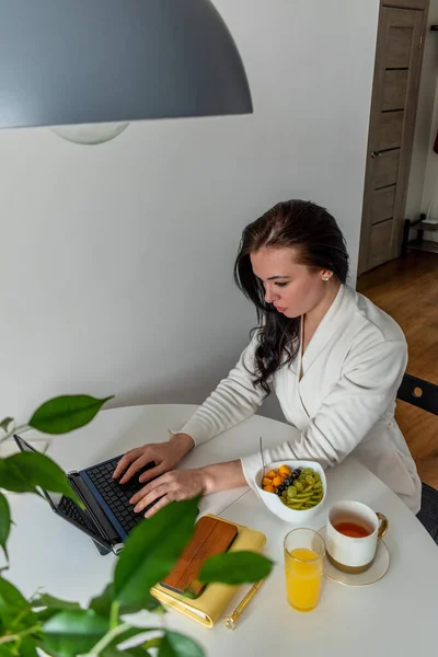 Young woman in comfortable white robe works on laptop while sitting at ...