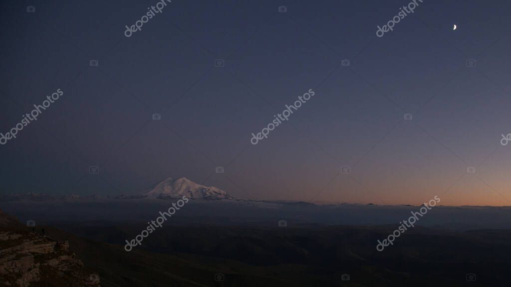 Elbrus en los últimos rayos del sol sobre el fondo de un cielo azul oscuro y una luna nueva ...