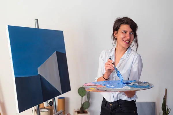 A young smiling painter mixes colors on a palette in her home studio ...