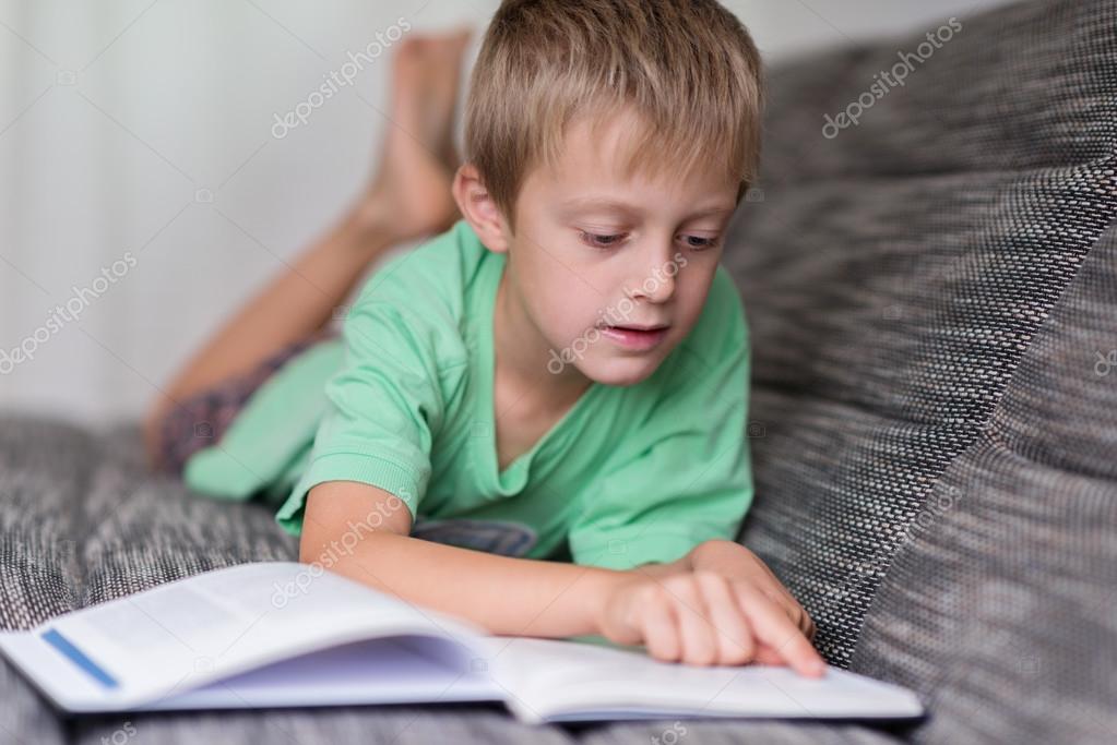 Niño pequeño acostado en un sofá leyendo un libro grande 2023