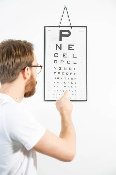 A young bearded man reading an eye test chart on a white wall. An ...