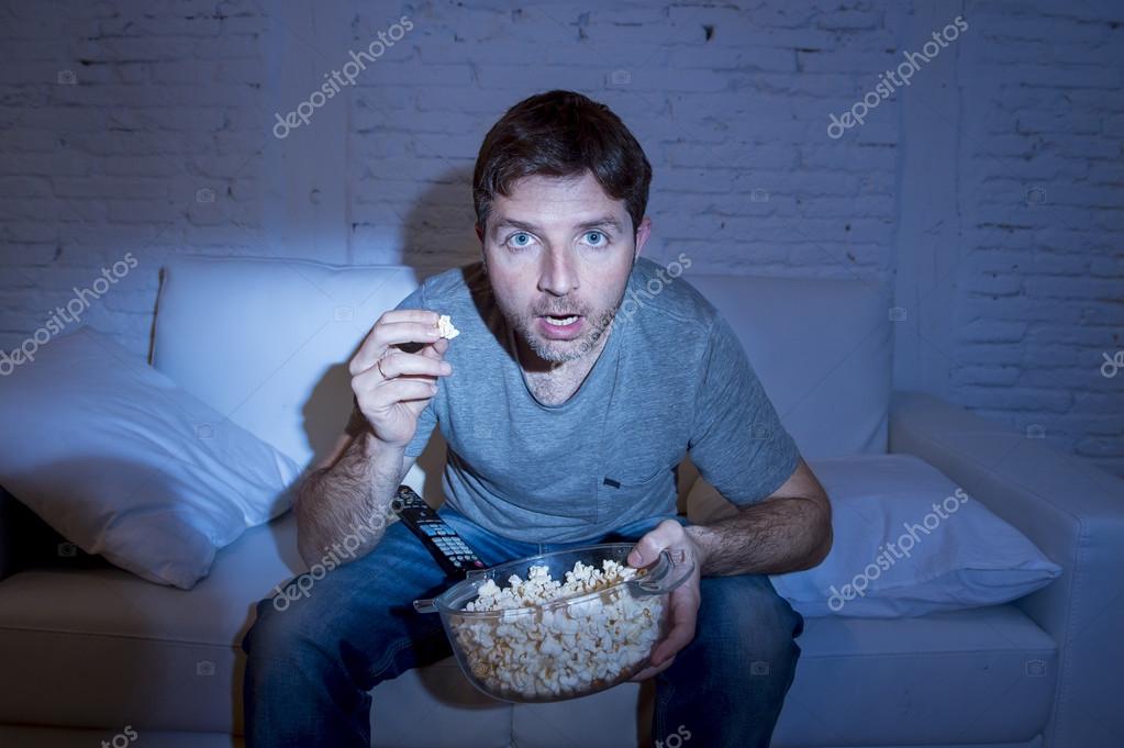 Young attractive man at home lying on couch watching tv holding popcorn ...