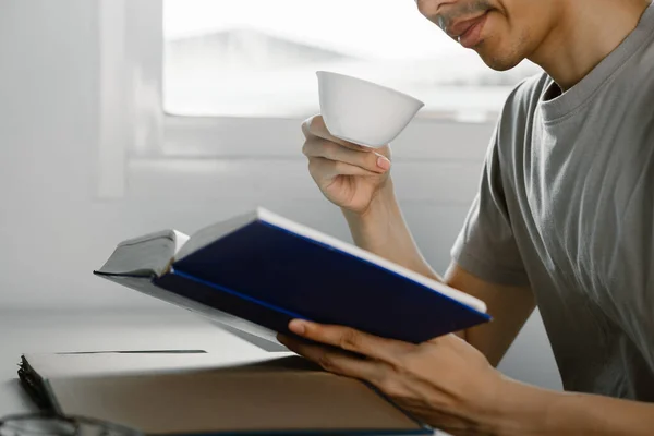 Young man reading book and drinking tea at work desk in free time from ...