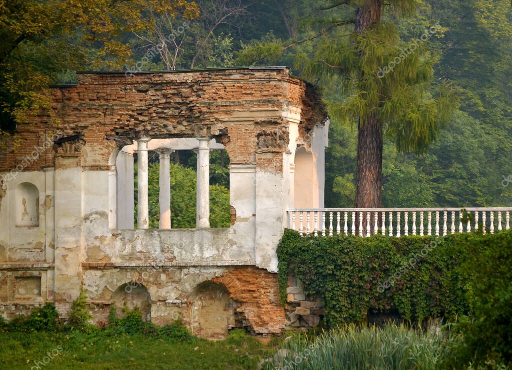 Ruins of red brick with white railing with columns and plaster in the park with green trees in ...