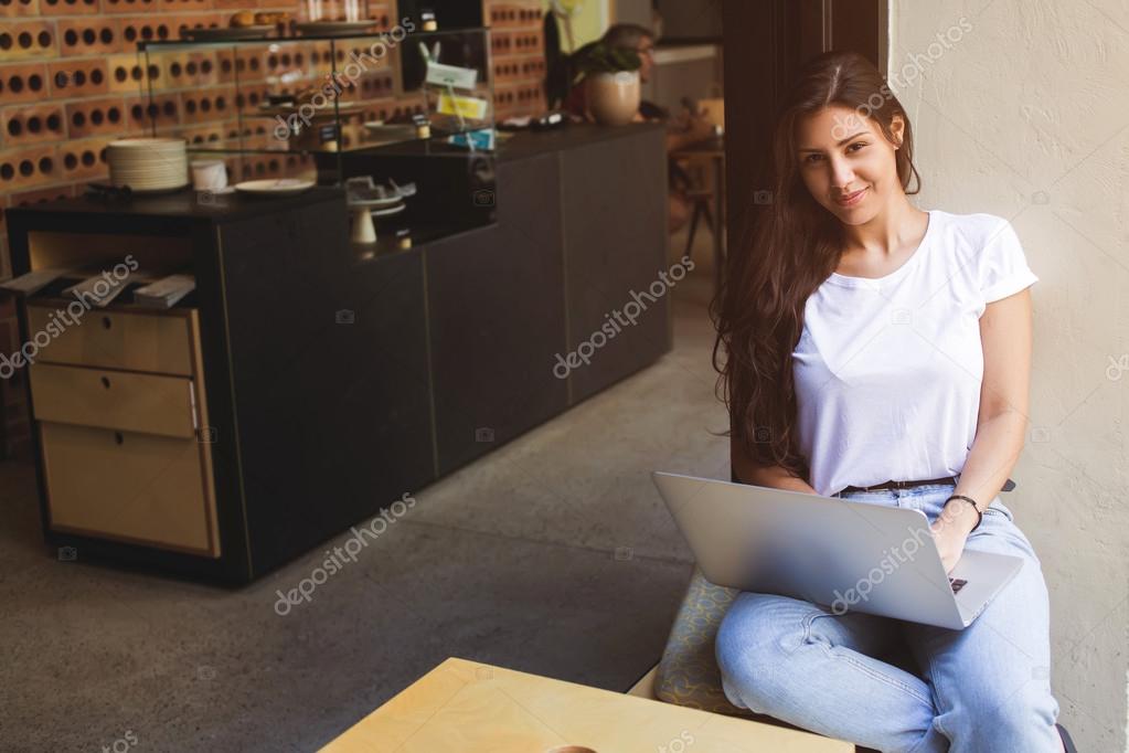 Gorgeous female freelancer using net-book for distance job while sitting in modern coffee shop ...
