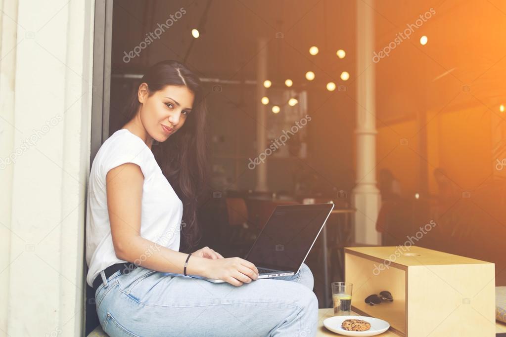 Female freelancer is using net-book for remote job while sitting in modern sidewalk coffee shop ...