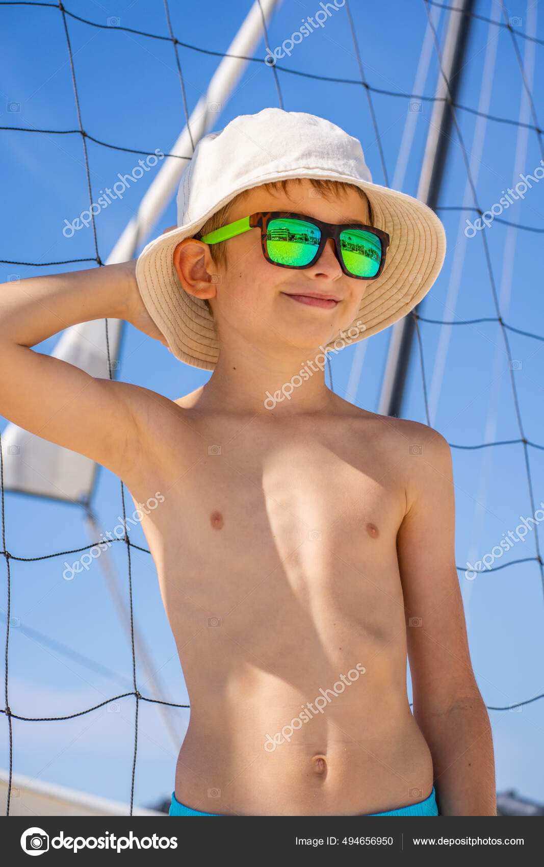 Handsome young boy in blue shorts, sunglasses and hat standing on white sand on a volleyball ...
