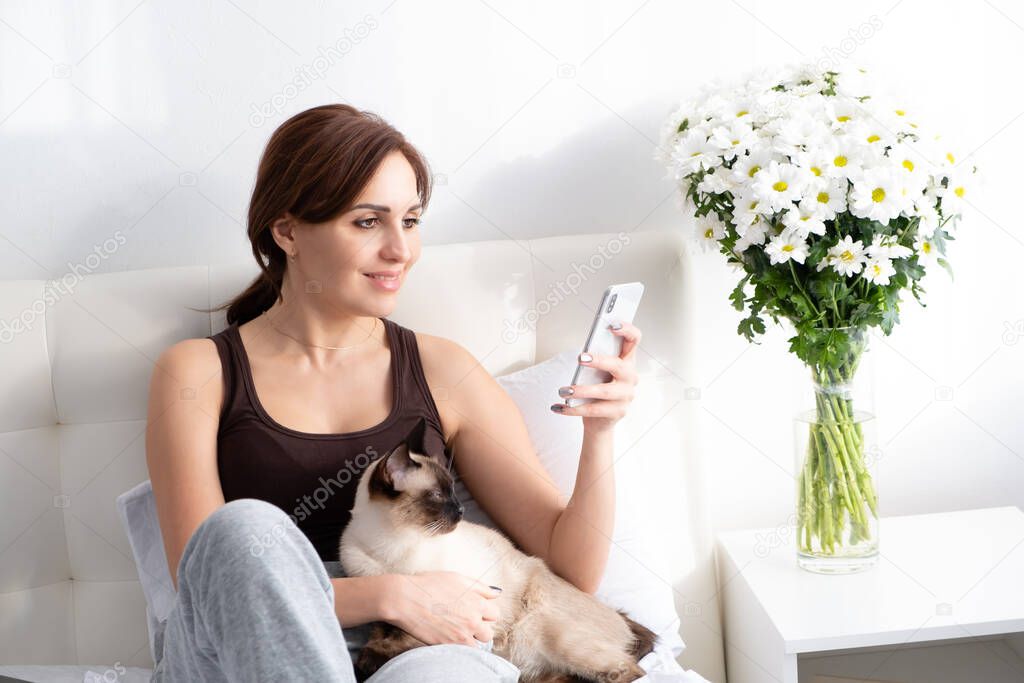 mujer joven usando el teléfono con su gato siamés en el dormitorio con ...