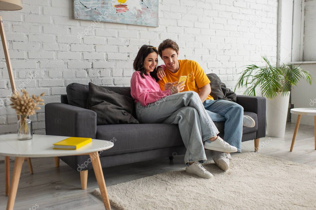pareja joven y positiva sentada en el sof y sosteniendo el tel fono celular en la sala de estar 2024