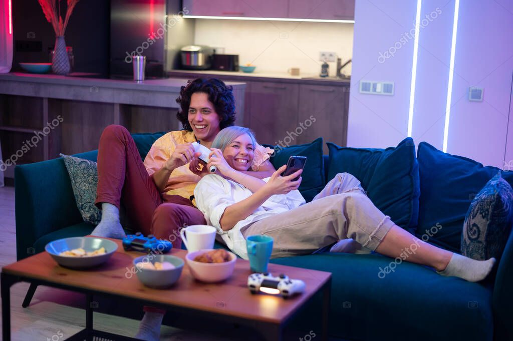 Feliz novio y novia jugando videojuegos y sosteniendo joysticks en casa. Pareja milenaria ...